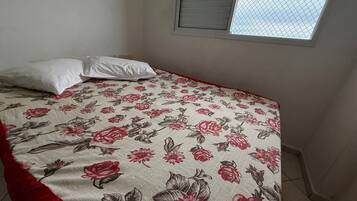 Uma cama com colcha florida, um travesseiro branco e uma janela com mosquiteiro.