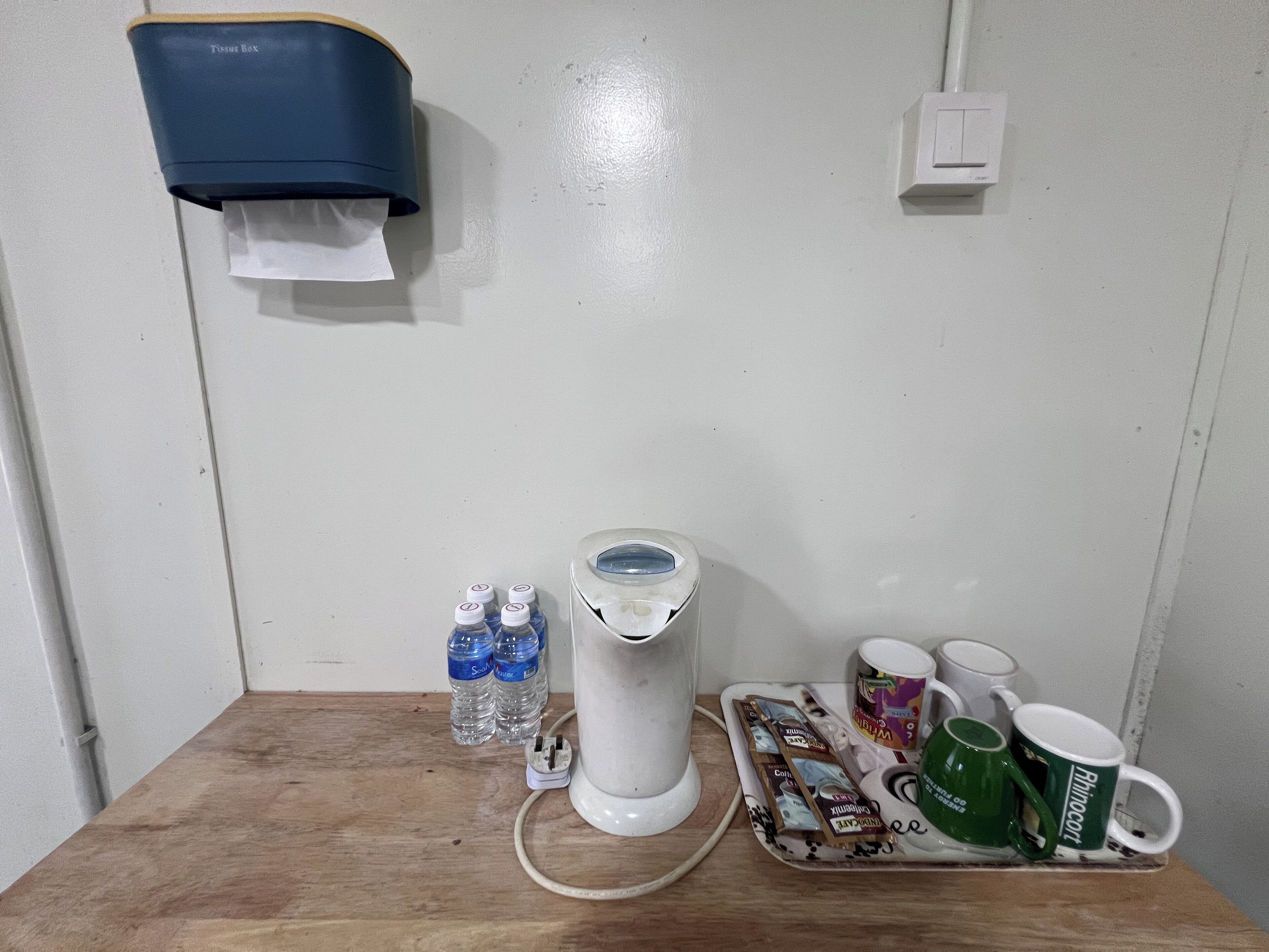 Uma pequena cozinha compacta com cafeteira, garrafas de água, caixa de lenços de papel e bandeja com cápsulas de café e canecas.