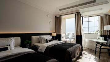 Quarto de hotel com duas camas, uma escrivaninha, uma cadeira e vista para o mar.