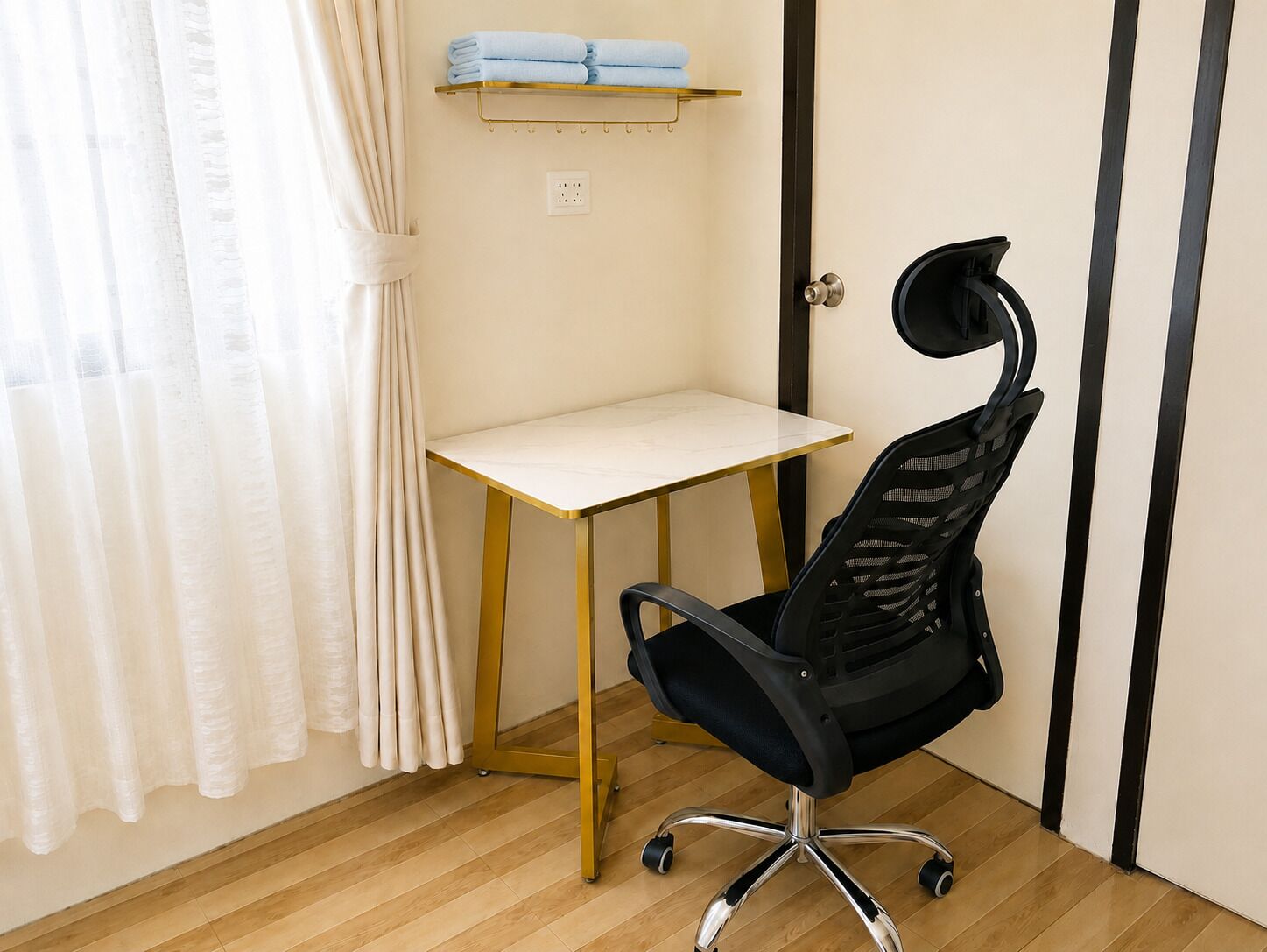Um pequeno espaço de escritório com uma mesa branca, uma cadeira de escritório preta, uma escrivaninha com moldura dourada e uma janela com cortinas brancas.