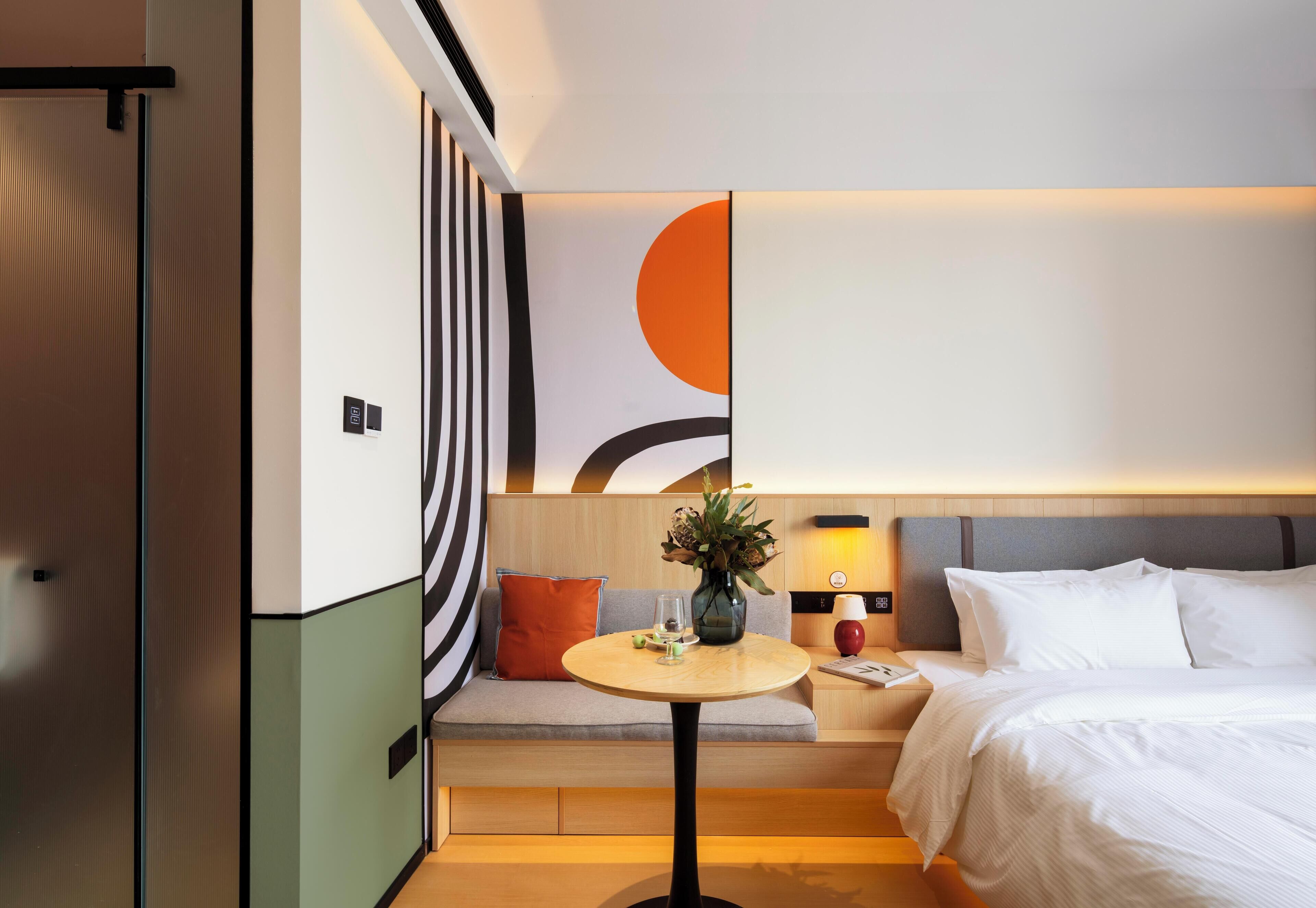 Quarto de hotel moderno com uma cama, uma mesa redonda com um vaso, uma mesinha de cabeceira e uma luminária de parede.