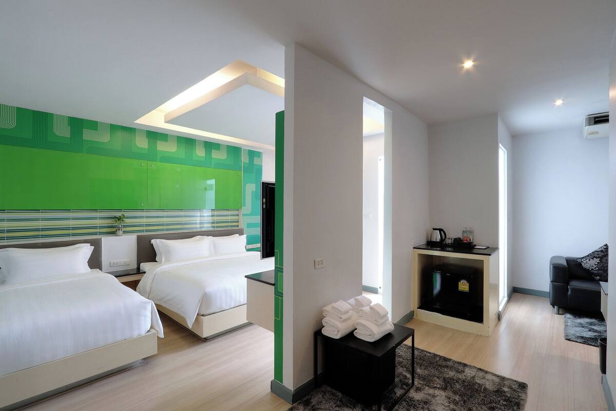 Quarto de hotel moderno com duas camas, uma parede verde de destaque, uma pequena cozinha e uma área de estar com um sofá preto.