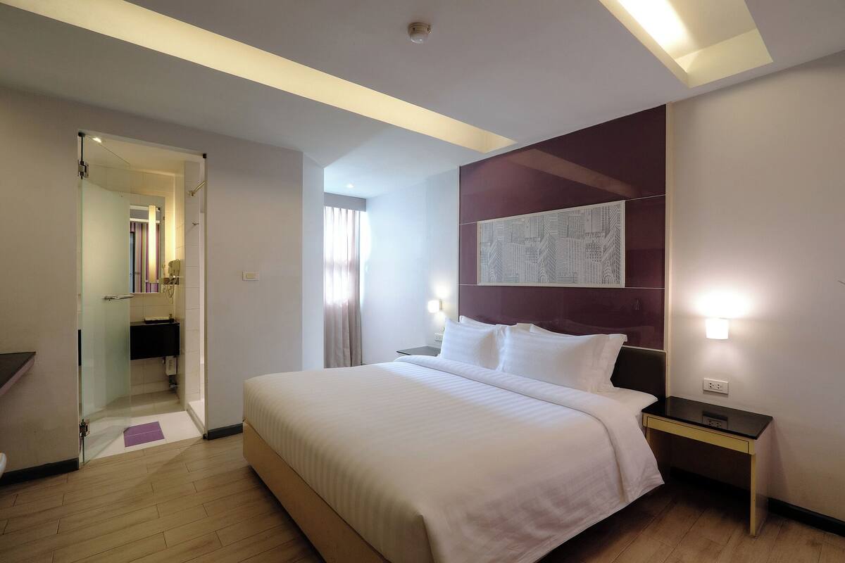 Quarto de hotel com uma cama grande, mesa de cabeceira, obra de arte na parede e banheiro visível através de uma porta aberta.