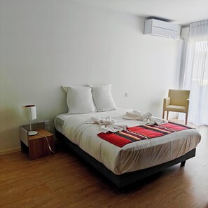 Uma cama bem arrumada com lençóis brancos e um cobertor listrado em vermelho, uma mesa de cabeceira com abajur, e uma cadeira junto à janela.