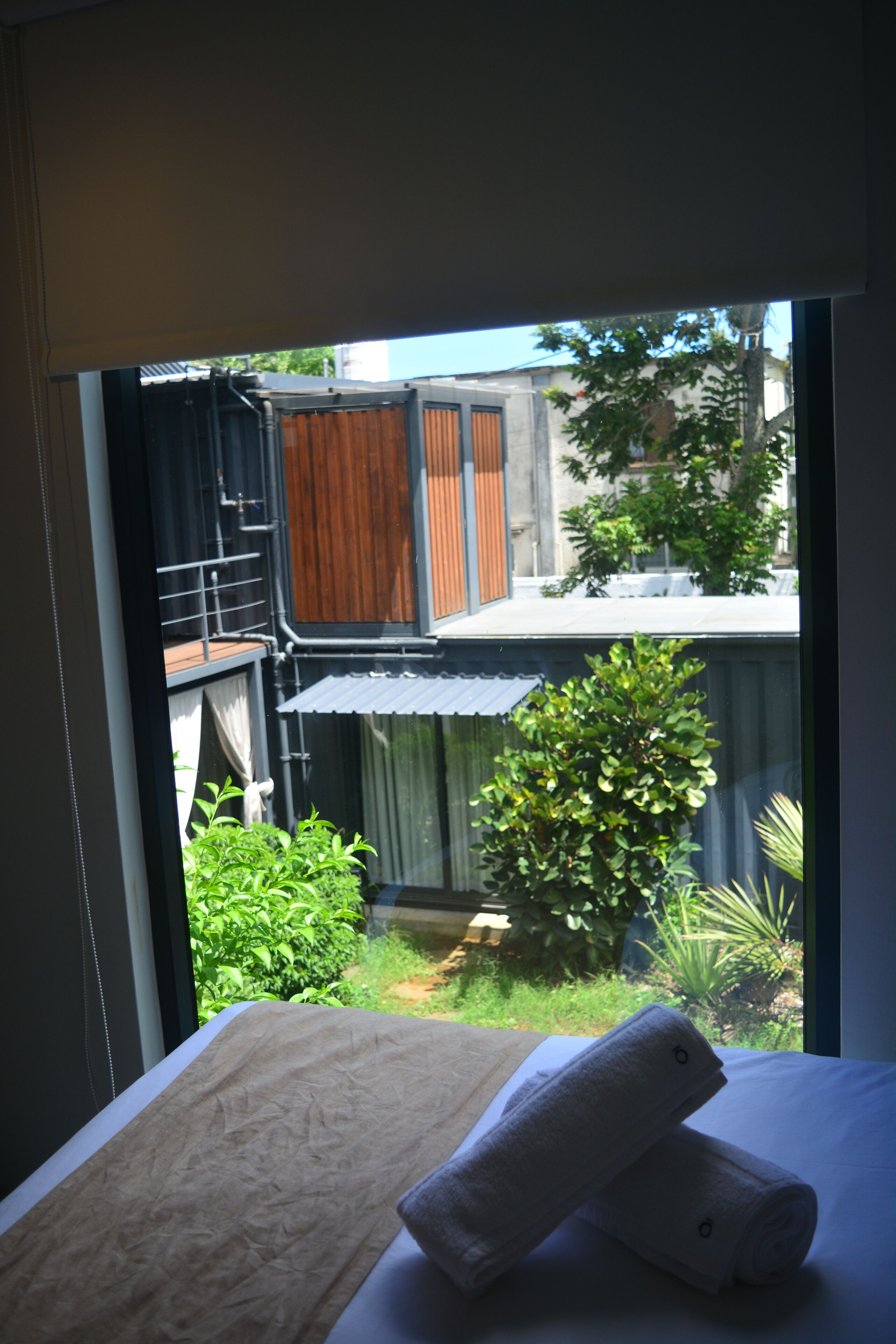 Cama com toalha branca e vista para um pátio com vegetação e um edifício.