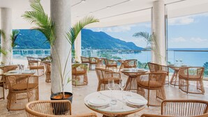 Restaurante com móveis de vime e vista para o mar e as montanhas.