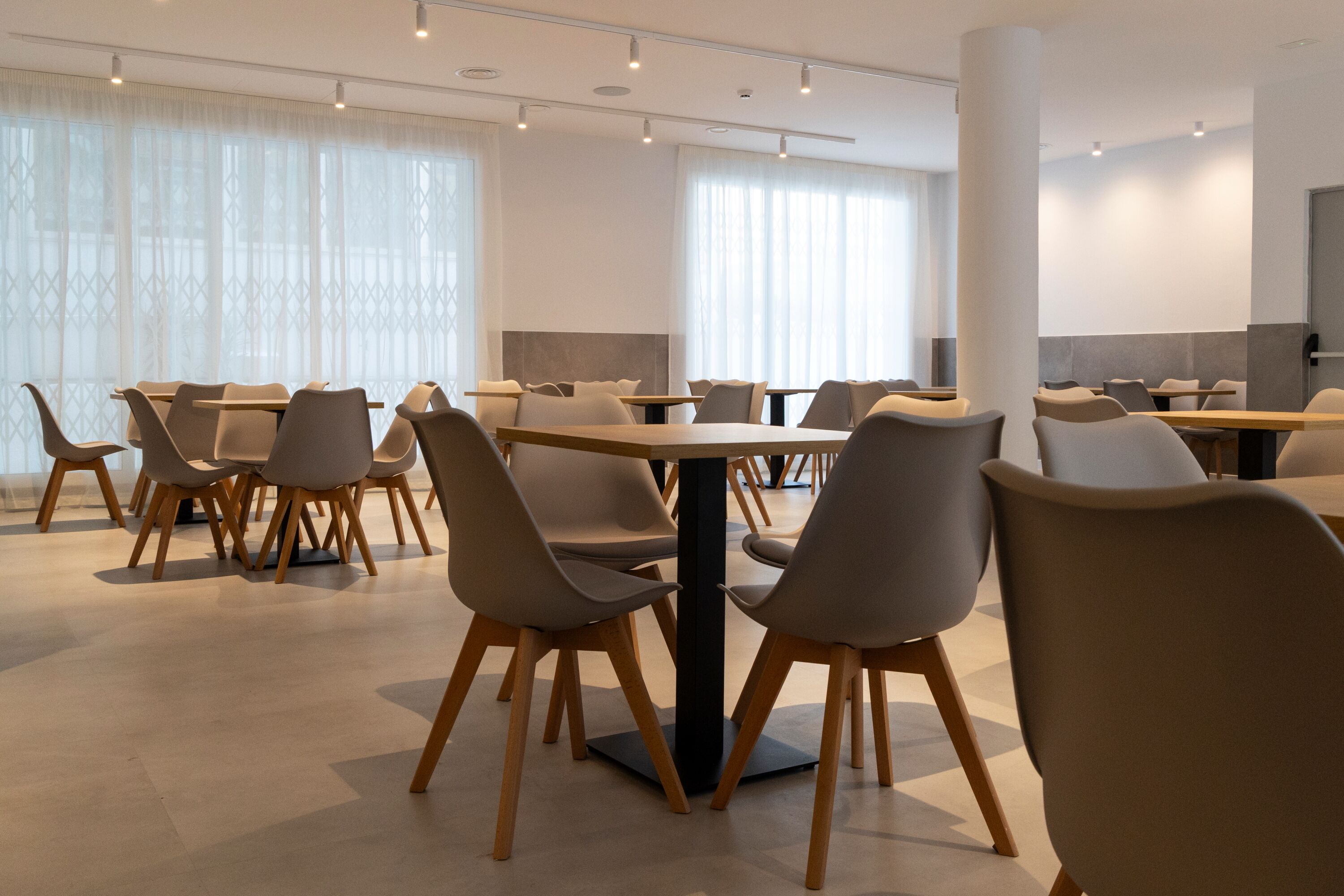 Uma sala de jantar moderna com mesas e cadeiras redondas, grandes janelas com cortinas leves e iluminação embutida no teto.