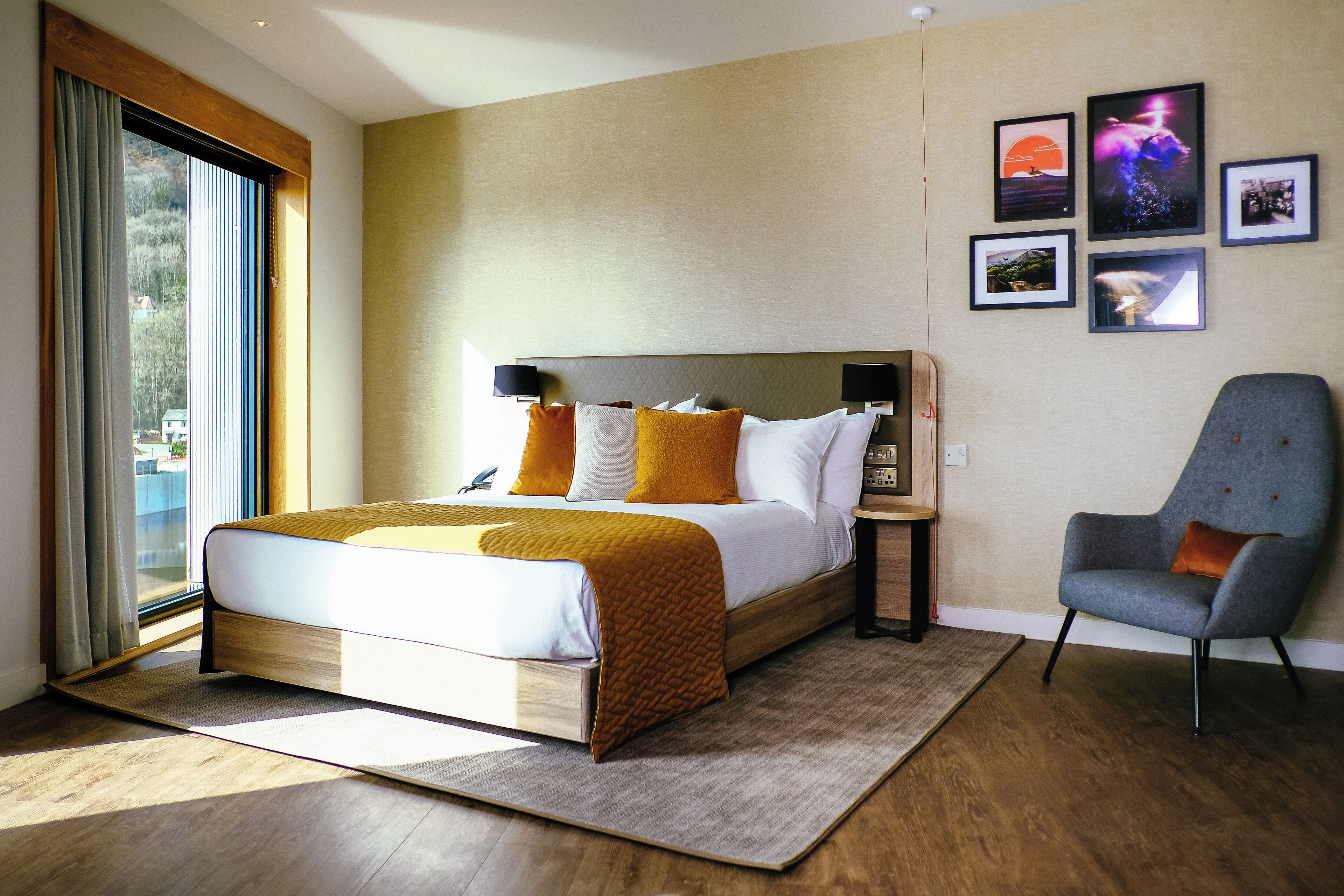 Quarto de hotel moderno com uma cama grande, uma mesinha de cabeceira, uma cadeira e vista para o exterior.