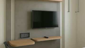 Quarto de hotel moderno com televisão de tela plana presa a uma parede cinza, prateleiras de madeira, uma mesinha e uma cadeira preta.
