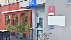 Entrada de hotel com um banner azul com a inscrição "ibis budget hotels", uma placa vermelha com um 'H' branco e estrelas, e uma porta com painel de vidro.