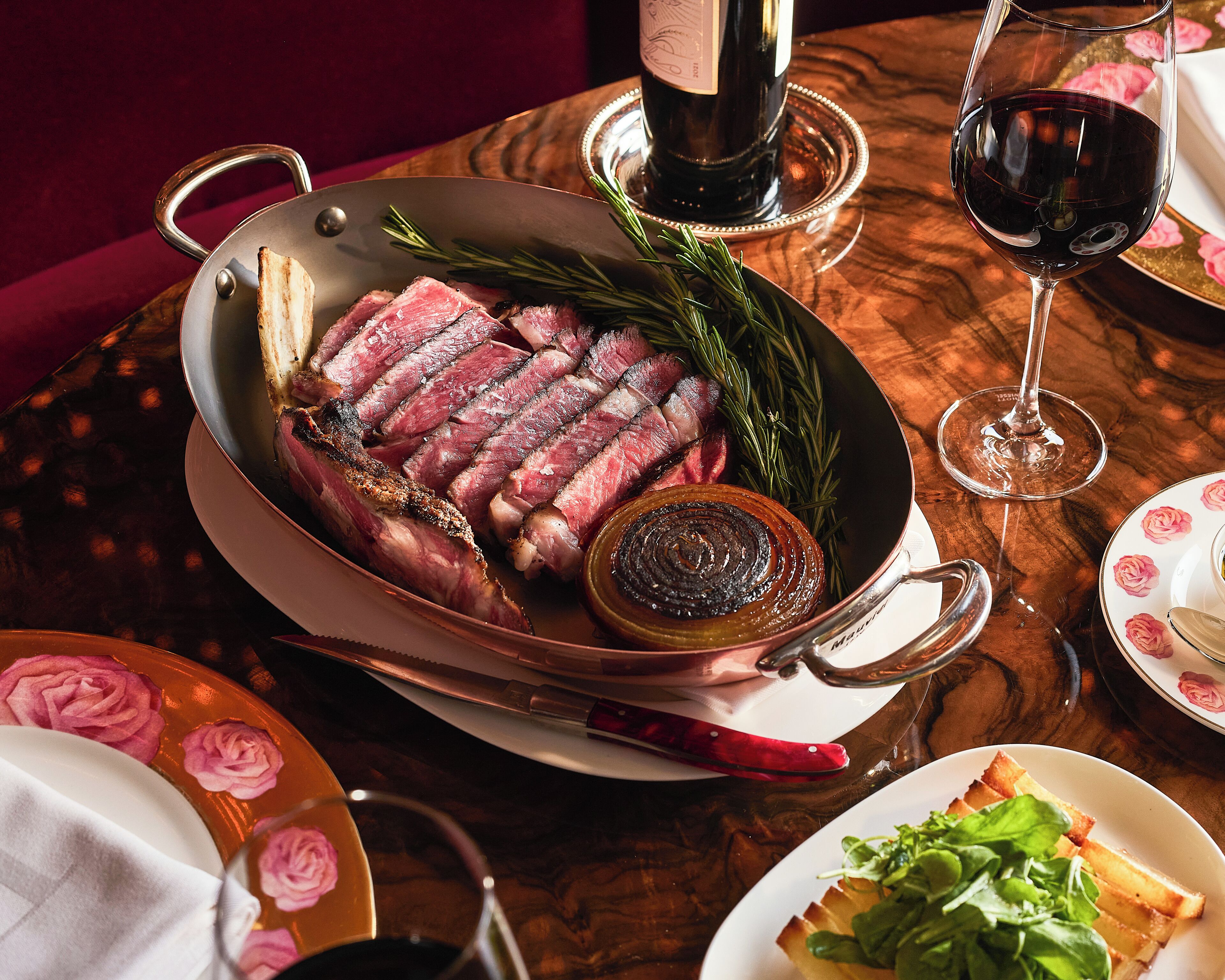 Uma frigideira com bife fatiado, alecrim e um acompanhamento de molho, acompanhados por um copo de vinho tinto e um prato de folhas verdes.