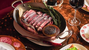 Uma frigideira com bife fatiado, alecrim e um acompanhamento de molho, acompanhados por um copo de vinho tinto e um prato de folhas verdes.