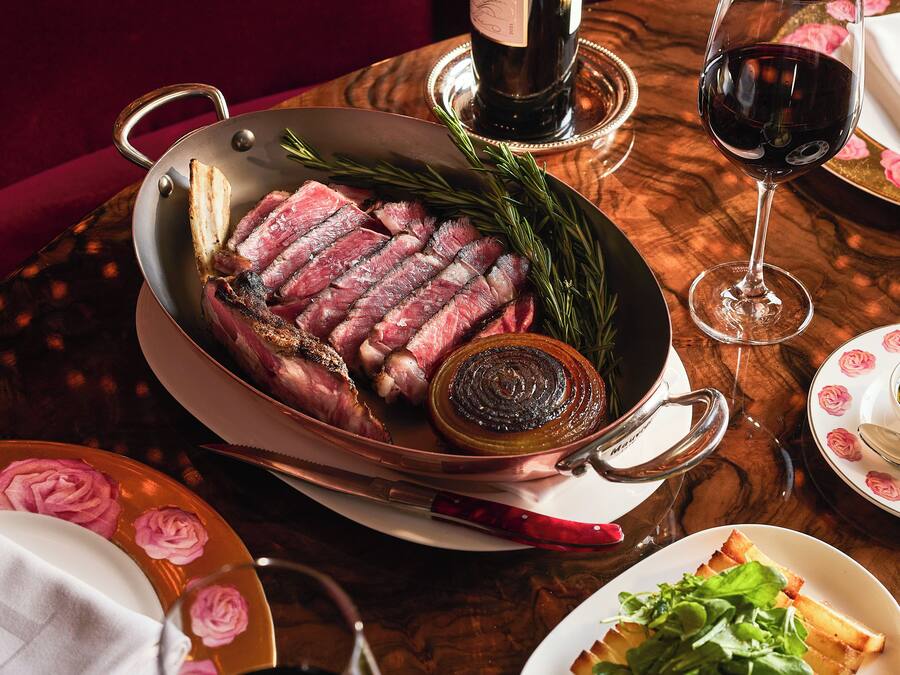 Uma frigideira com bife fatiado, alecrim e um acompanhamento de molho, acompanhados por um copo de vinho tinto e um prato de folhas verdes.