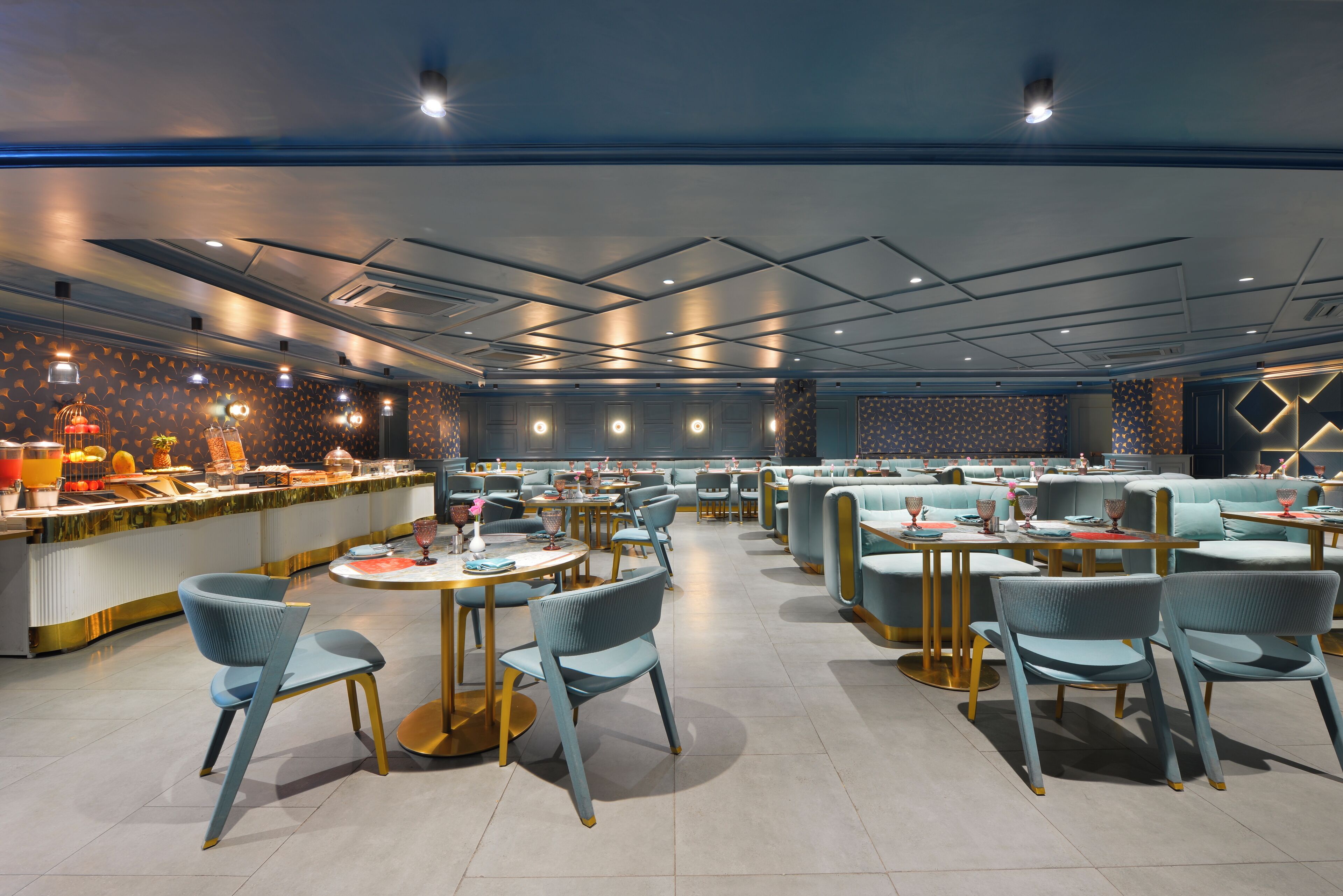 Um restaurante moderno com mesas redondas e cadeiras azuis, área de bar com exposição de frutas e teto com padrão decorativo.