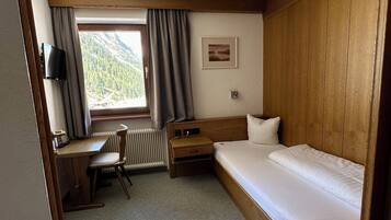 Quarto de hotel com cama de madeira, escrivaninha com cadeira, radiador, janela com cortinas e um quadro na parede.