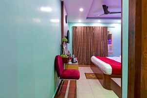 Quarto de hotel compacto com uma cama de solteiro, uma escrivaninha com cadeira, televisão e ventoinha de teto.