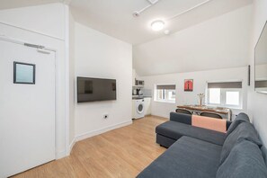 Uma sala de estar moderna com televisão de tela plana, sofás cinza, uma área de jantar com mesa, e uma cozinha ao fundo.