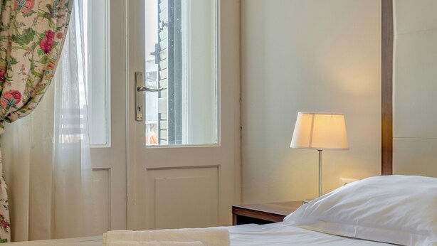 Quarto de hotel com cama, mesa de cabeceira, abajur e porta com janela.