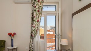 Quarto de hotel com cama, uma mesinha com um vaso de flores, uma cadeira, um abajur e vista para edifícios através de uma porta aberta.