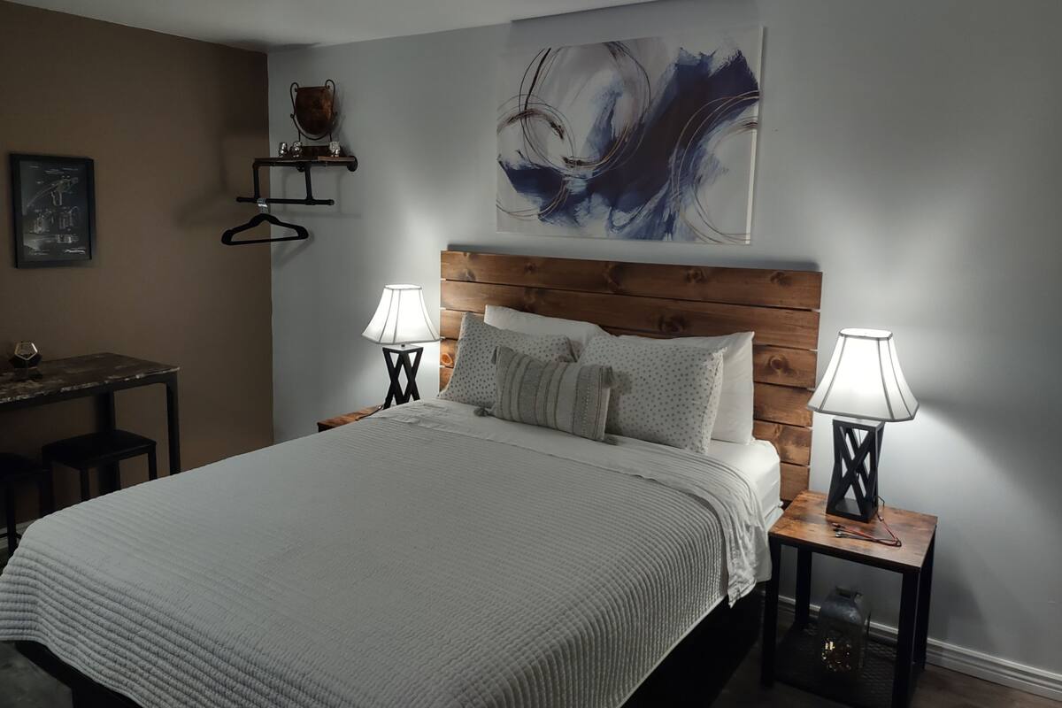 Quarto com uma cama grande, duas mesas de cabeceira com luminárias, cabeceira de madeira, prateleira na parede com um objeto decorativo e um quadro pendurado na parede.