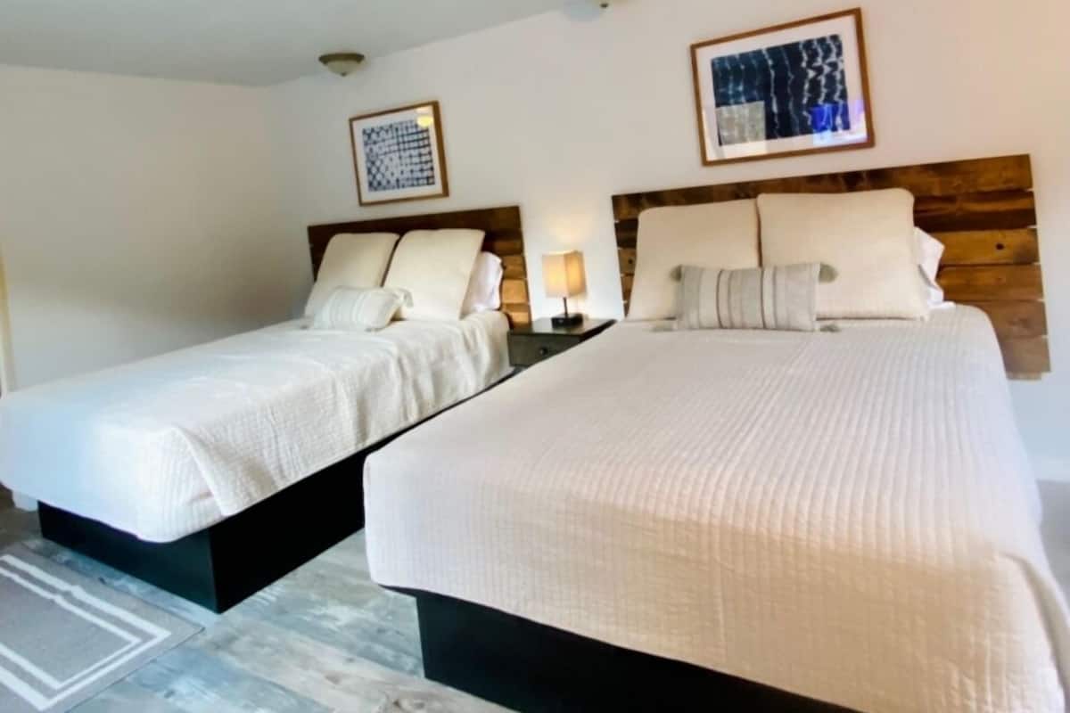 Quarto de hotel com duas camas, cada uma com cabeceira de madeira, roupa de cama branca e uma pequena mesa de cabeceira com abajur.