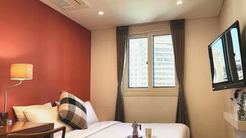 Quarto de hotel com cama, escrivaninha com luminária, televisão na parede e janela com cortinas.