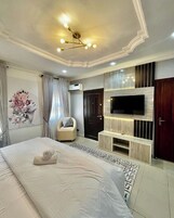 Quarto moderno com cama grande, televisão na parede, uma cadeira e um objeto decorativo na parede.