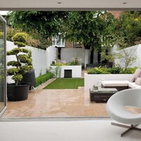 Uma área de estar ao ar livre moderna com paredes de vidro, um sofá branco, uma mesa de centro de vidro e um pequeno jardim com gramado e plantas em vasos.