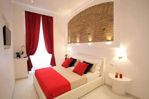 Quarto com cama grande, cortinas vermelhas, parede de tijolos em destaque e uma pequena mesa de cabeceira com duas velas vermelhas.