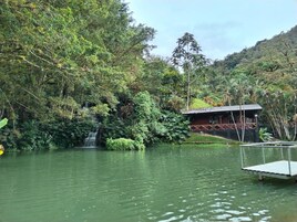 Um cenário tranquilo à beira de um lago, com um píer de madeira e um pequeno edifício, cercado por vegetação exuberante e uma cachoeira.