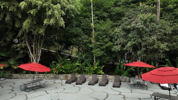 Uma área à beira da piscina com espreguiçadeiras, guarda-sóis vermelhos e um deck com ladrilhos de pedra.