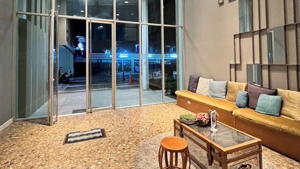Uma sala de estar moderna com uma grande porta de vidro que dá para uma área de estacionamento, um sofá com almofadas, uma mesa de centro e um banquinho.