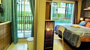 Quarto com cama, mesa de cabeceira, janela com cortinas, um banquinho pequeno e vista para o exterior.