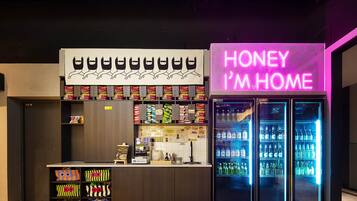 Uma área de bar com um letreiro de neon que diz 'HONEY I'M HOME', prateleiras repletas de lanches e uma geladeira cheia de bebidas.
