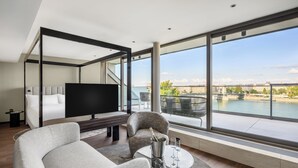Uma sala de estar moderna com uma televisão grande, assentos confortáveis, uma mesa de vidro com taças de vinho e vista para um rio e uma ponte.