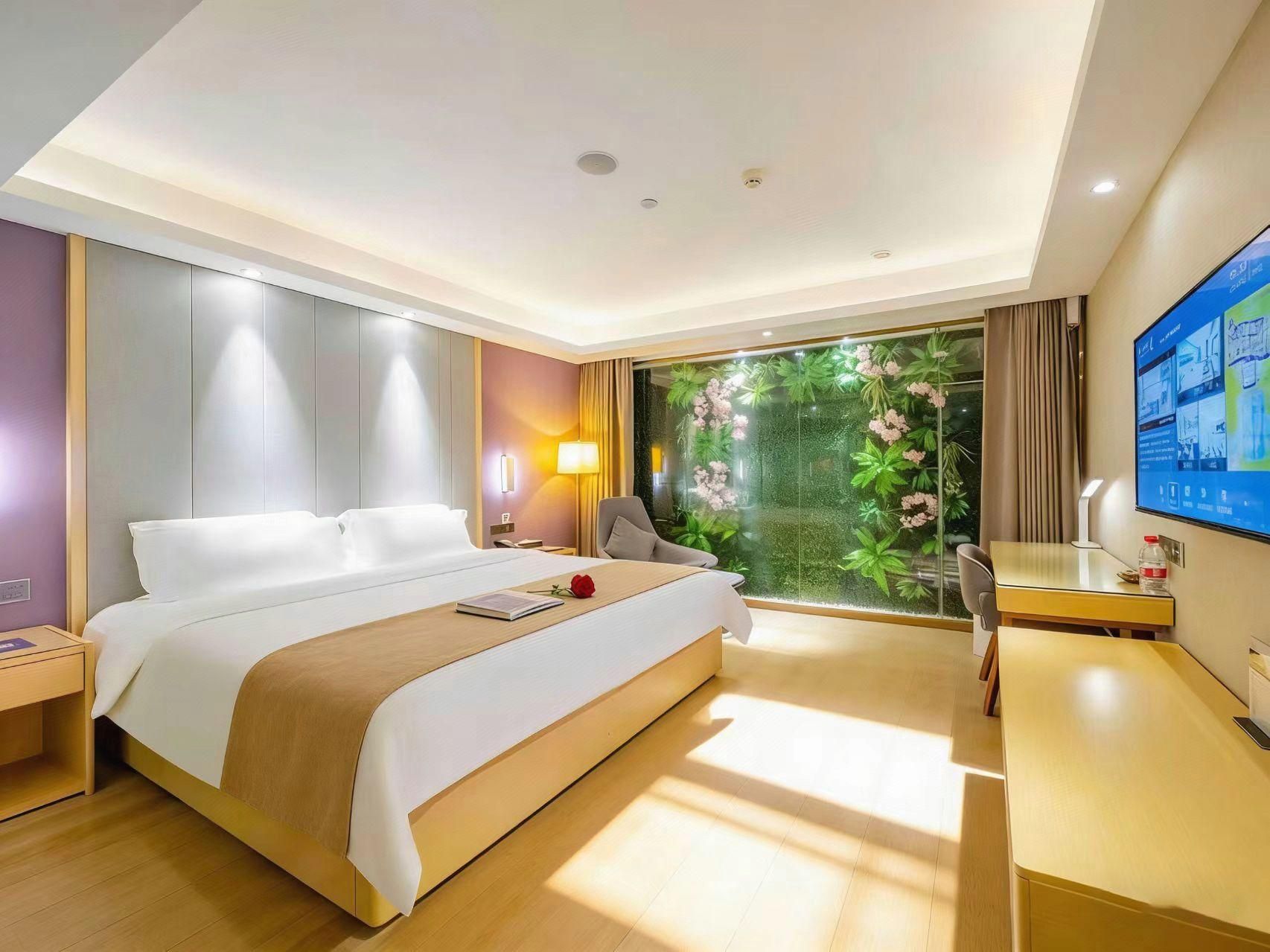 Quarto de hotel moderno com cama, escrivaninha, cadeira, televisão e uma grande janela com vista para o verde.