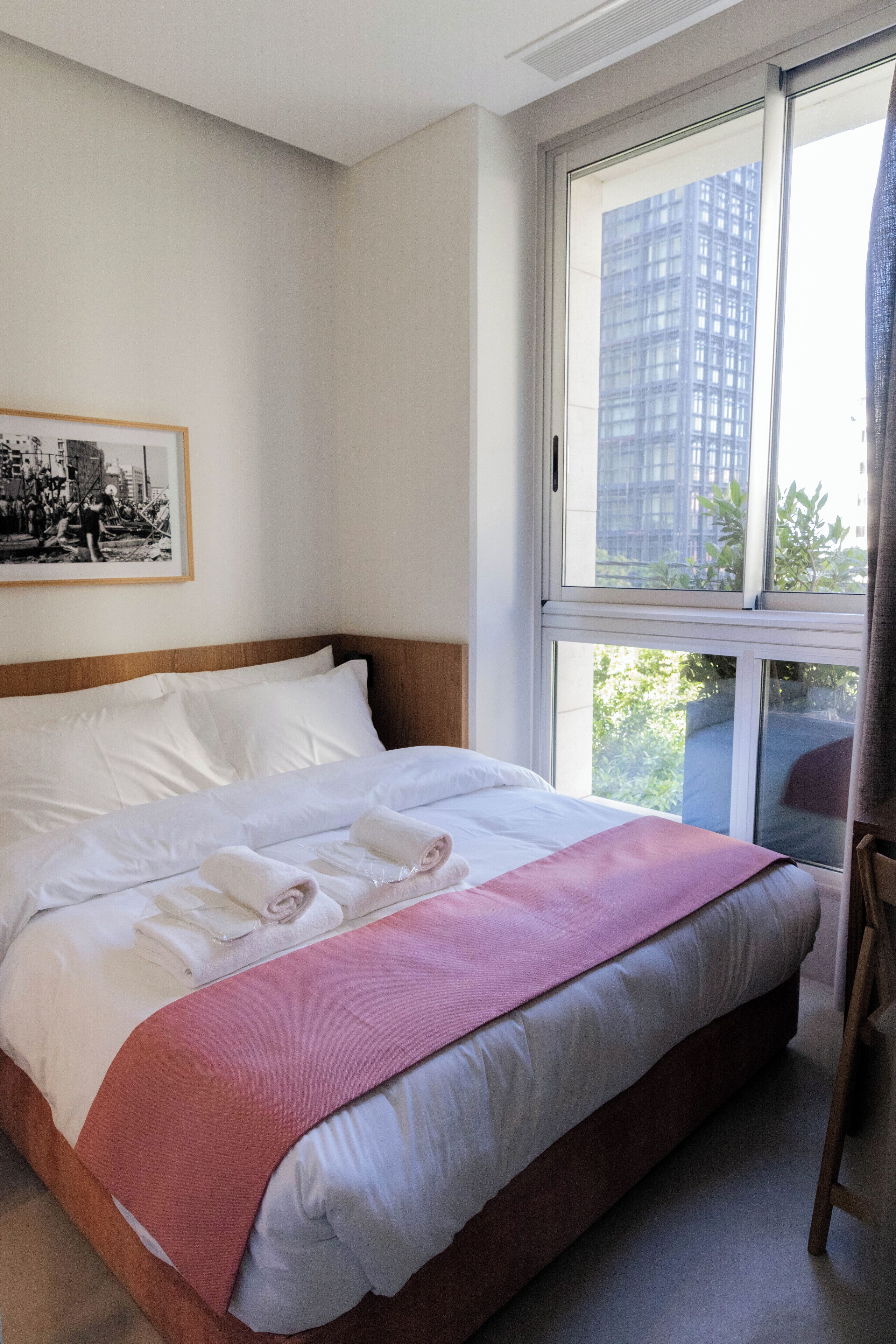 Cama bem arrumada com roupa de cama branca e rosa, um quadro na parede e uma janela com vista para edifícios e áreas verdes.