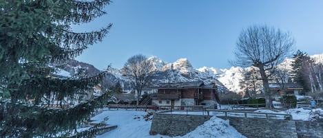 Paisagem coberta de neve com um prédio de madeira, um muro de pedra e montanhas nevadas ao fundo.