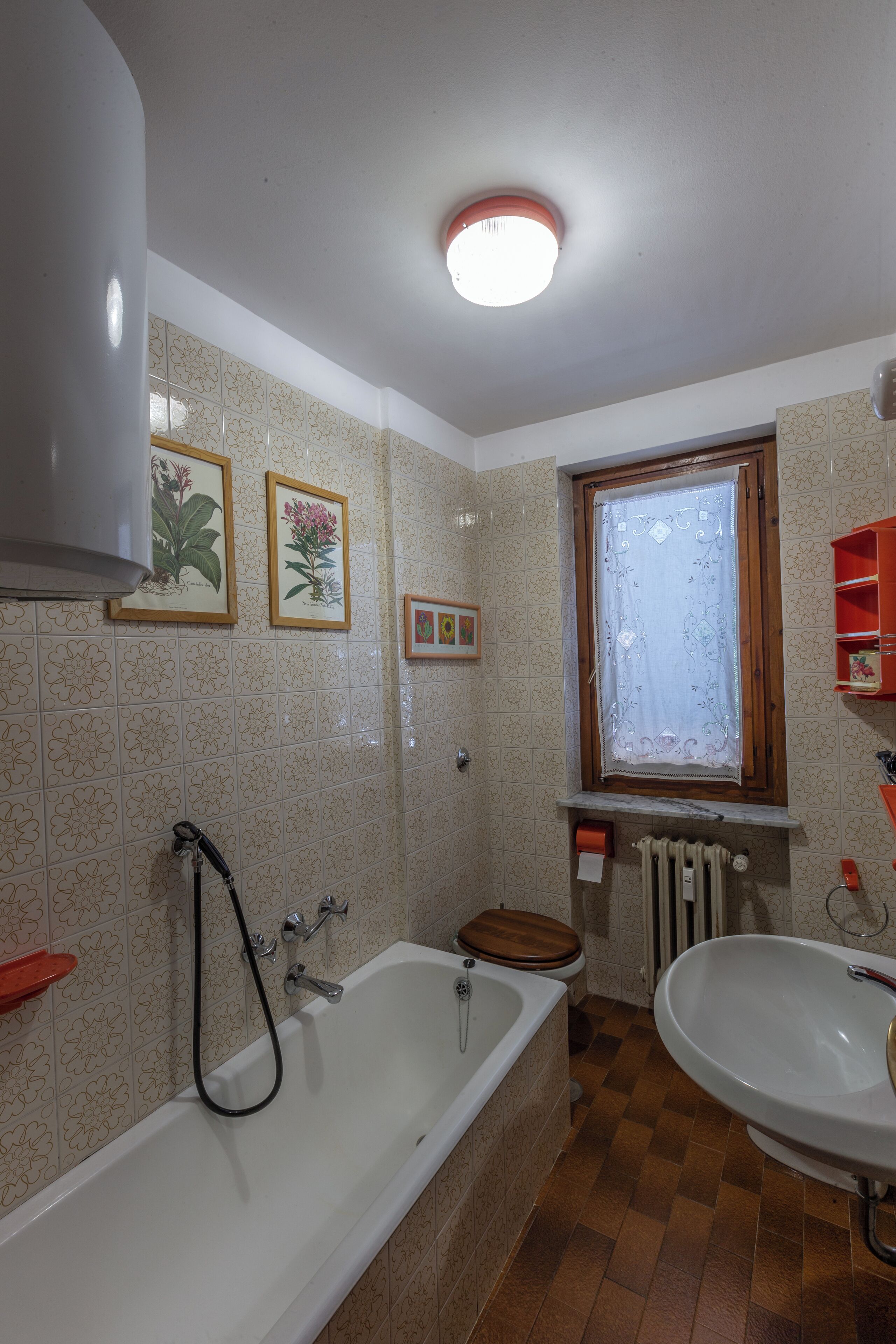 Casa de banho com banheira, lavatório e sanita. Apresenta azulejos com motivos florais e uma janela com cortina.