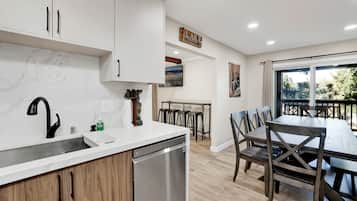 Cozinha moderna com armários brancos, lava-louças e painel de parede em mármore. Há uma área de refeições com mesa e cadeiras de madeira, e uma placa com a inscrição 'LAKE'.