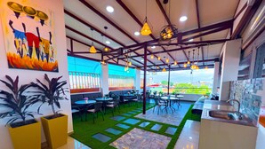 Um restaurante moderno com mesas ao ar livre, grama sintética verde, piso de pedra e luminárias pendentes.