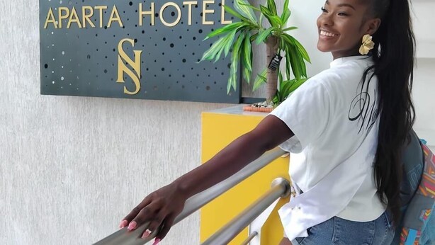 Uma pessoa apoiada num corrimão de metal em frente a um letreiro com a inscrição 'APARTA HOTEL', com um grande logotipo com as letras 'S' e 'N'.