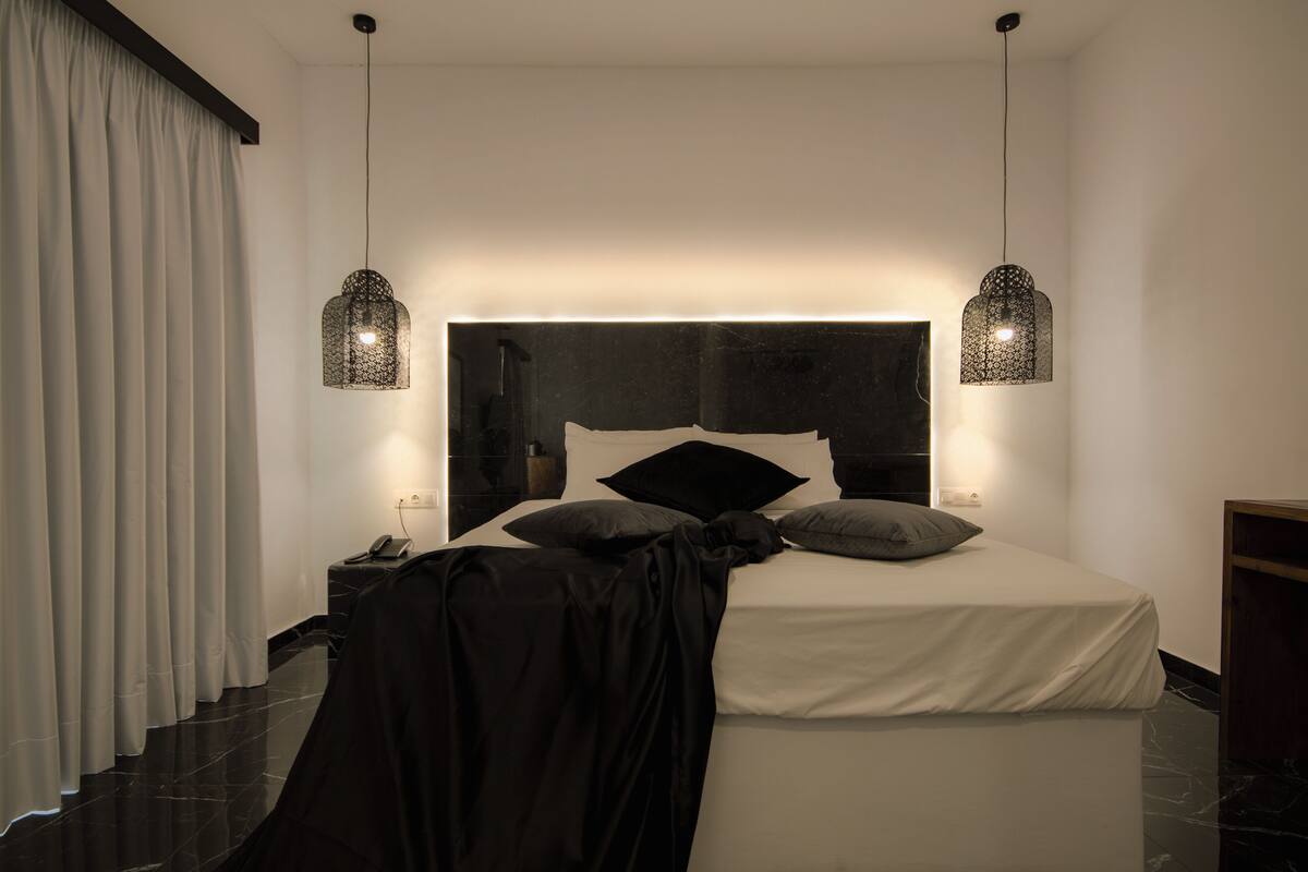 Quarto moderno com uma cama grande, duas luminárias pendentes, uma mesinha de cabeceira e uma cabeceira escura.