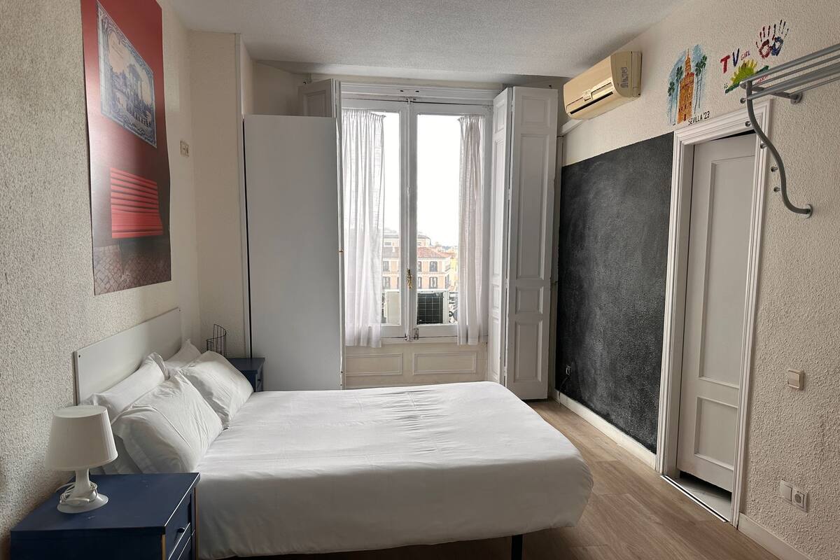 Quarto com cama, mesa de cabeceira, abajur, guarda-roupa e janela com cortinas.