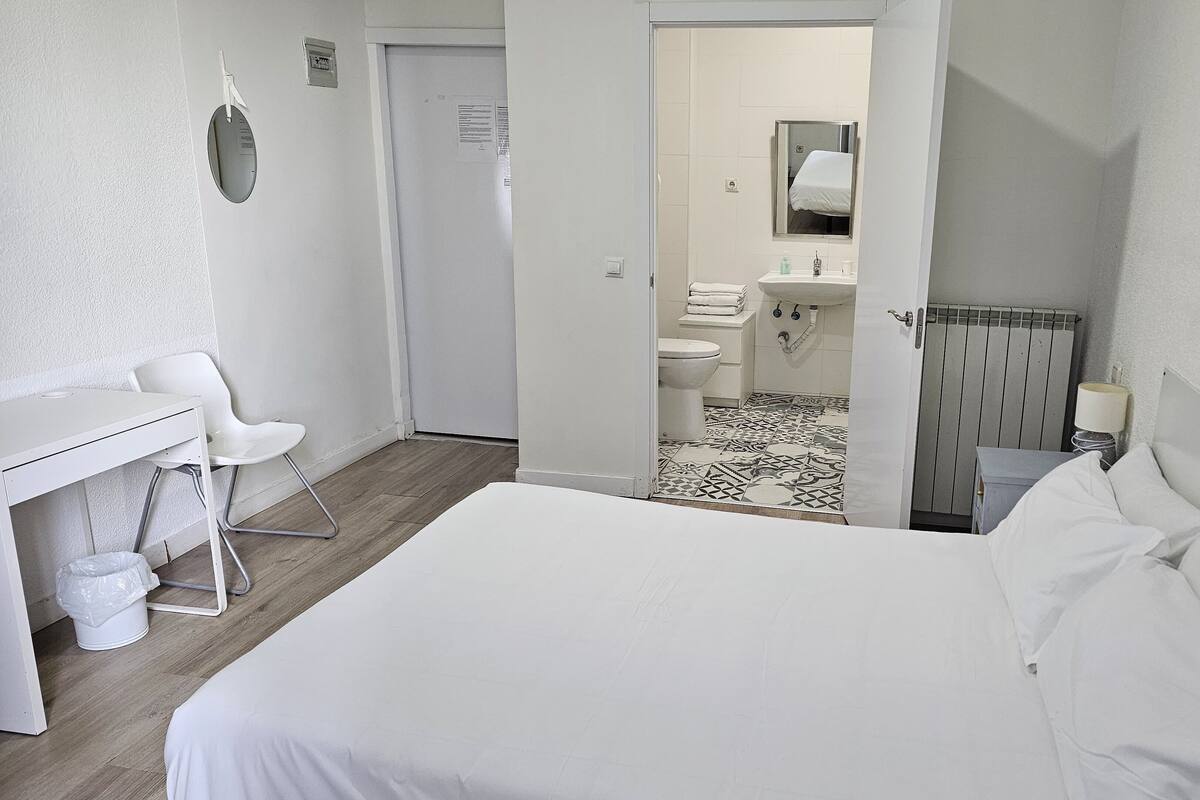 Quarto de hotel com cama, escrivaninha, cadeira e banheiro completo com vaso sanitário, pia e espelho.