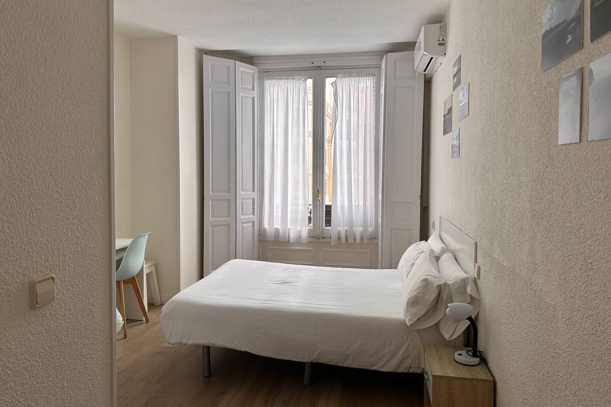 Quarto com cama, mesa de cabeceira e abajur, uma cadeira e vista pela janela com cortinas.