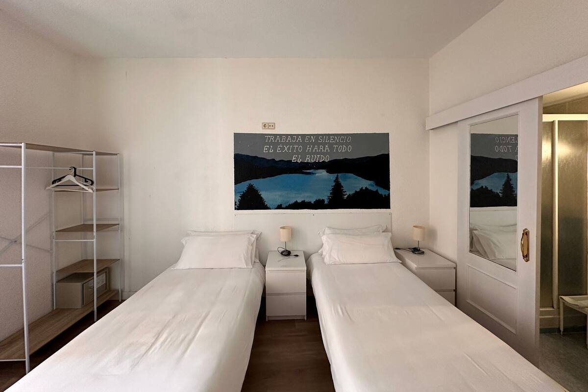 Quarto de hotel com duas camas de solteiro, um guarda-roupa branco, uma mesa de cabeceira com uma luminária e um pôster na parede.