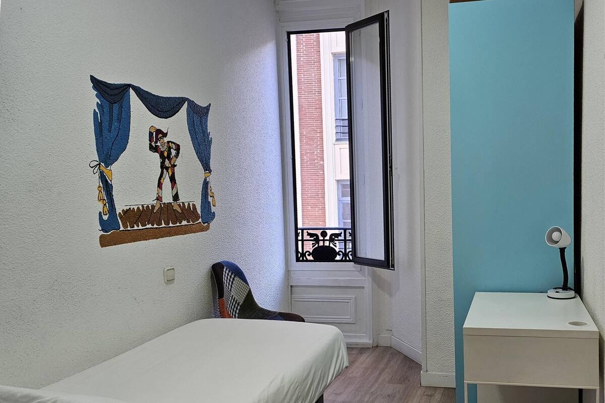 Quarto com cama, escrivaninha, cadeira, janela com vista para um edifício e parede com um mural.