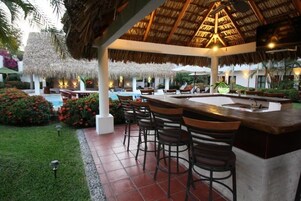 Uma área de bar ao ar livre com piscina, telhado de palha e banquetas.