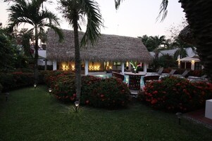 Um edifício com telhado de palha, piscina e espreguiçadeiras, rodeado por vegetação exuberante e flores vermelhas.